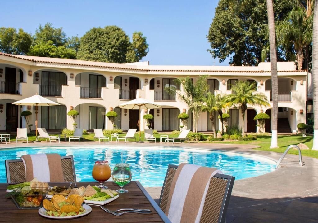 Uma área de refeições à beira da piscina, com uma mesa posta para dois, rodeada por espreguiçadeiras e guarda-sóis, e um edifício com varandas e piscina ao fundo.