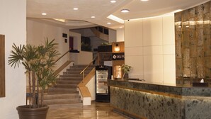 Um saguão de hotel com balcão de recepção, escada e uma grande planta em vaso.