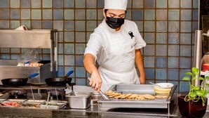 Cozinheiro com uniforme branco preparando comida em um buffet, com diversos alimentos e utensílios de cozinha.