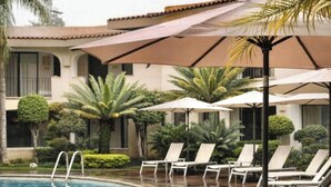 Piscina com espreguiçadeiras e guarda-sóis, cercada por palmeiras e um edifício com telhado de azulejos.