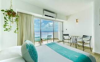 Quarto de hotel com uma cama grande, varanda com vista para o oceano, uma mesa pequena e cadeiras, e um ar condicionado.