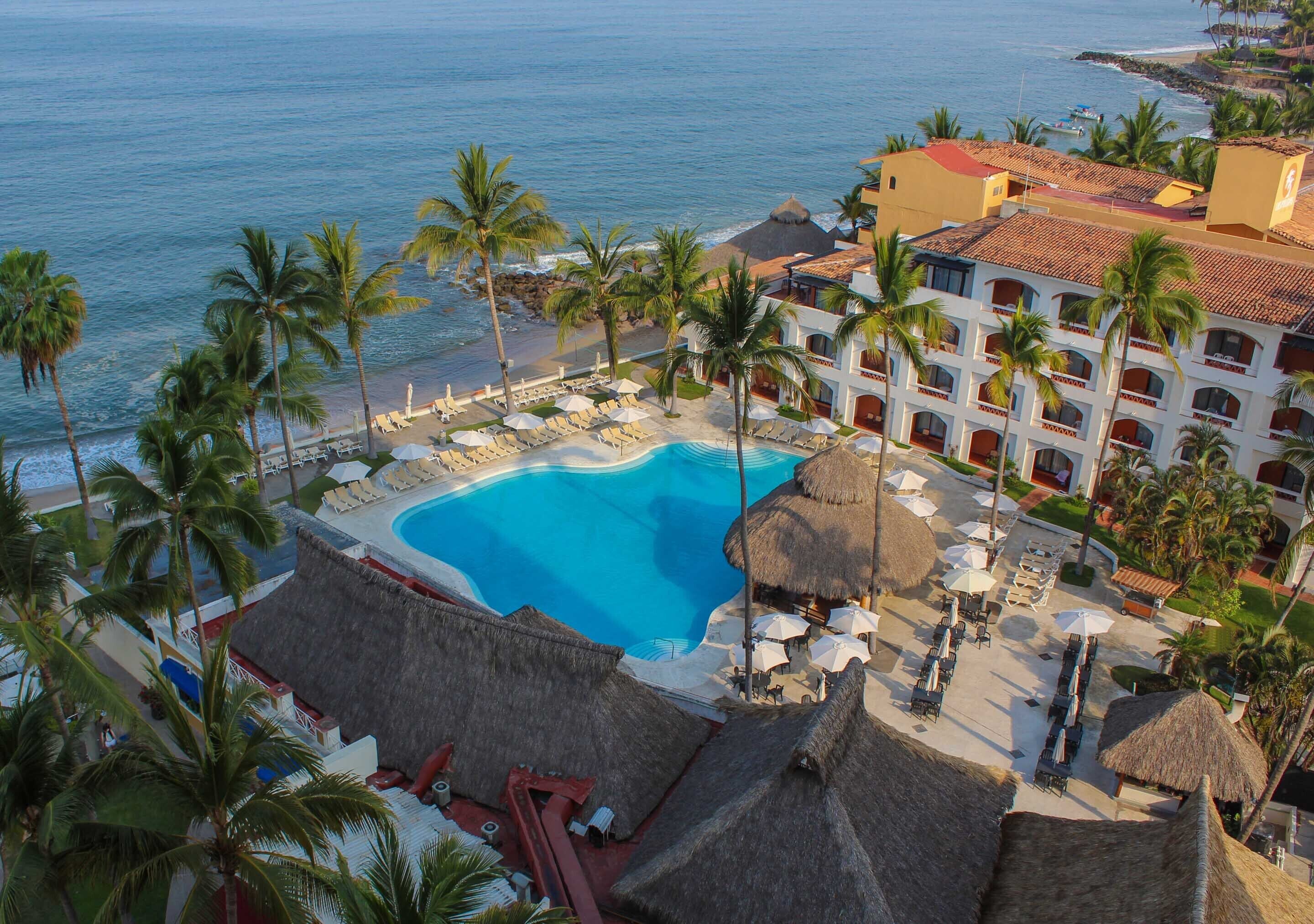 Resort com piscina, palmeiras e vista para a praia.