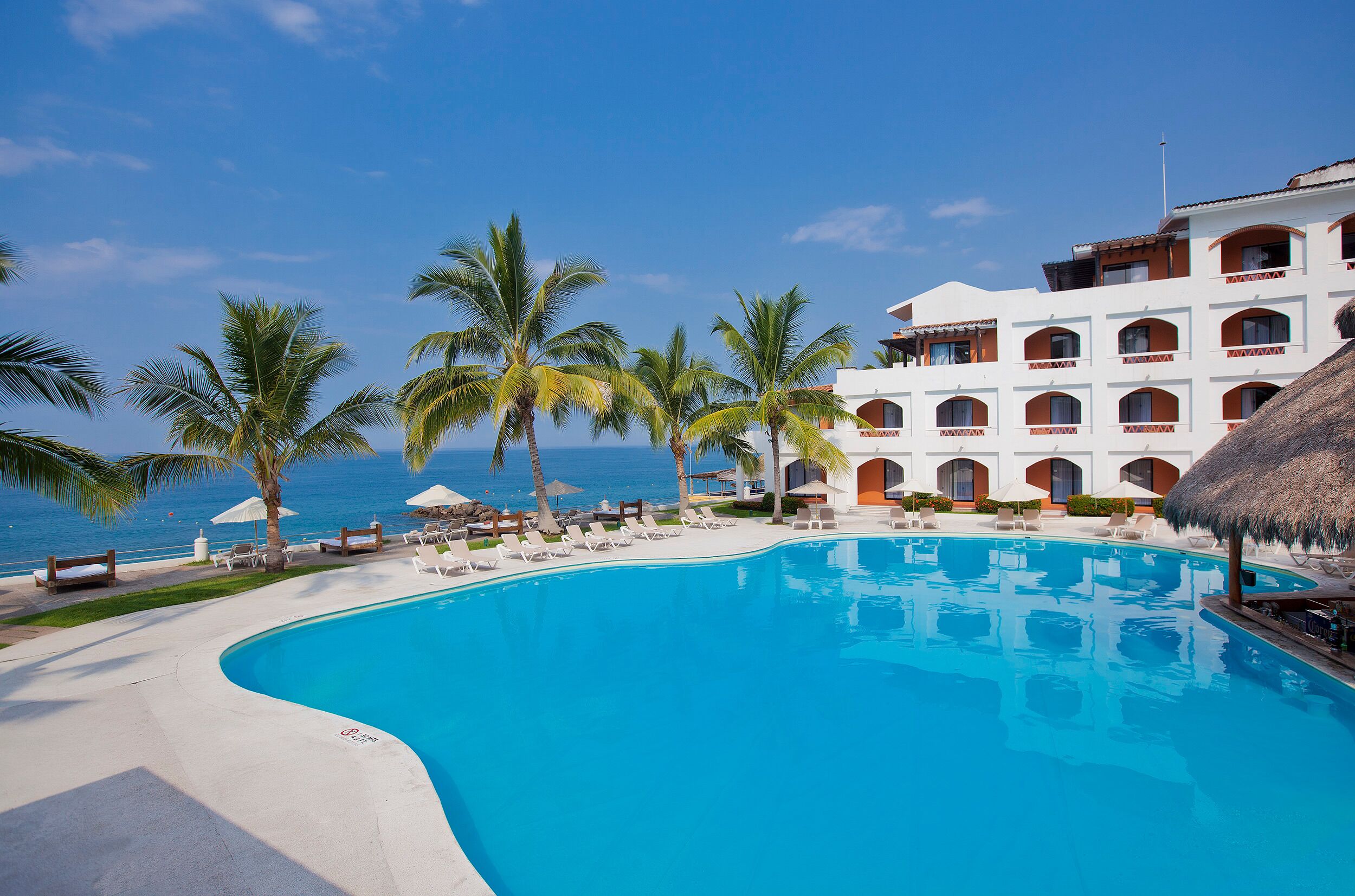 Piscina de resort com palmeiras, espreguiçadeiras e um edifício com varandas com vista para o mar.