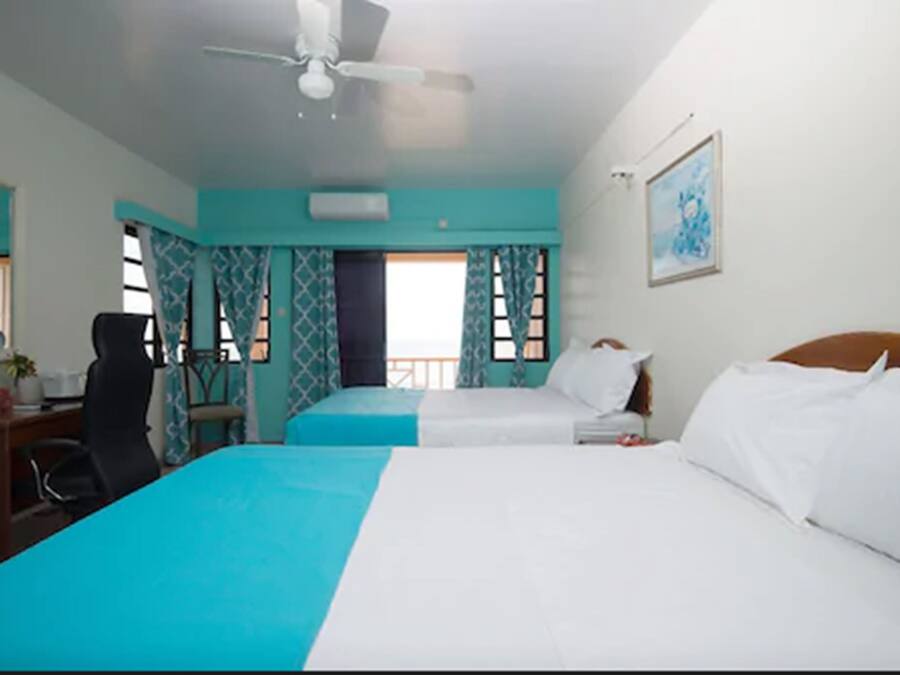 Quarto de hotel com duas camas, uma escrivaninha, uma cadeira e um ventilador de teto.
