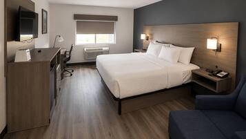 Quarto de hotel com cama, escrivaninha, cadeira, televisão, luminária e mesa de cabeceira com telefone.