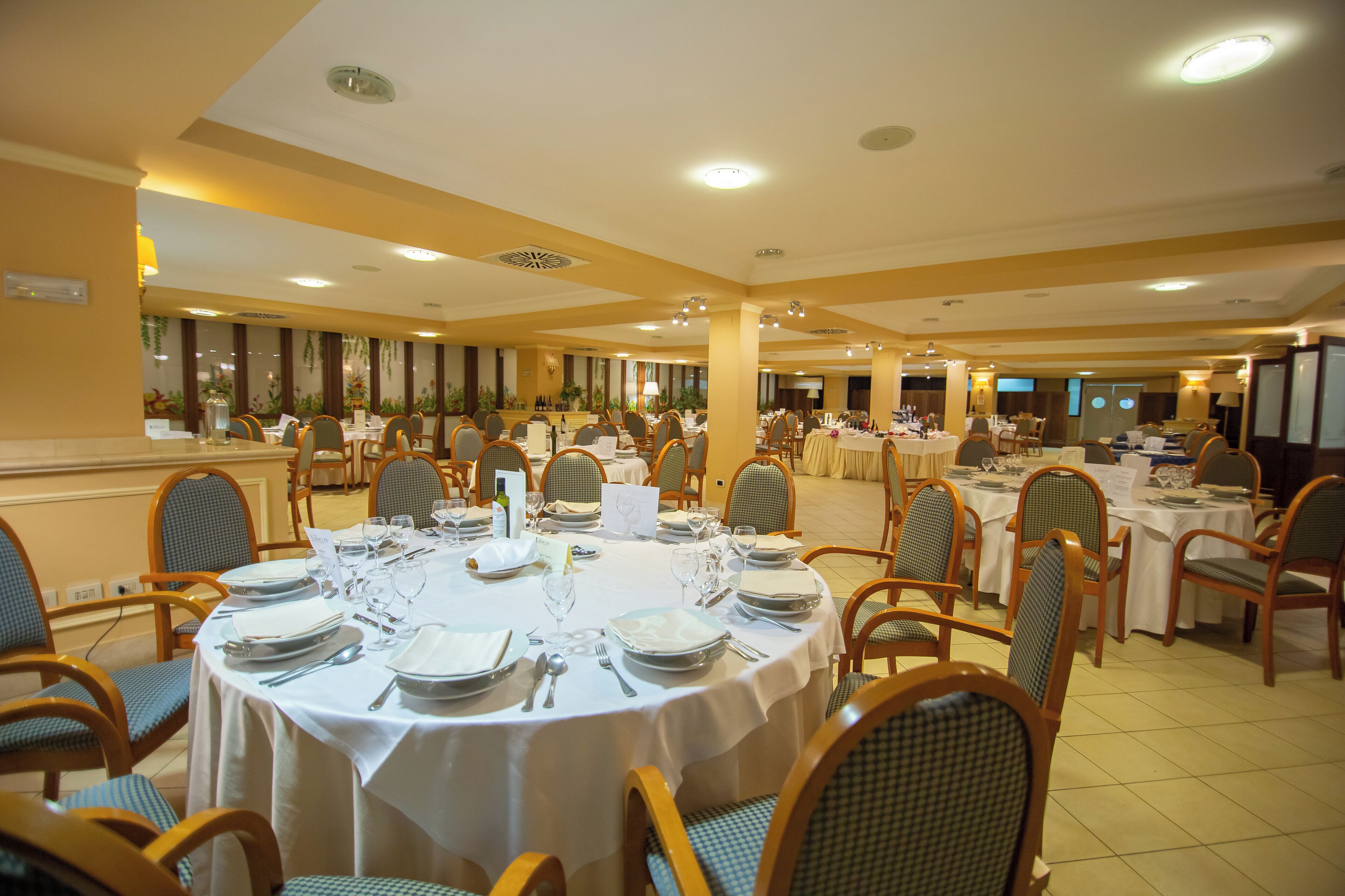 Um amplo salão de jantar com mesas redondas preparadas para vários convidados, com cadeiras de madeira e assentos acolchoados.