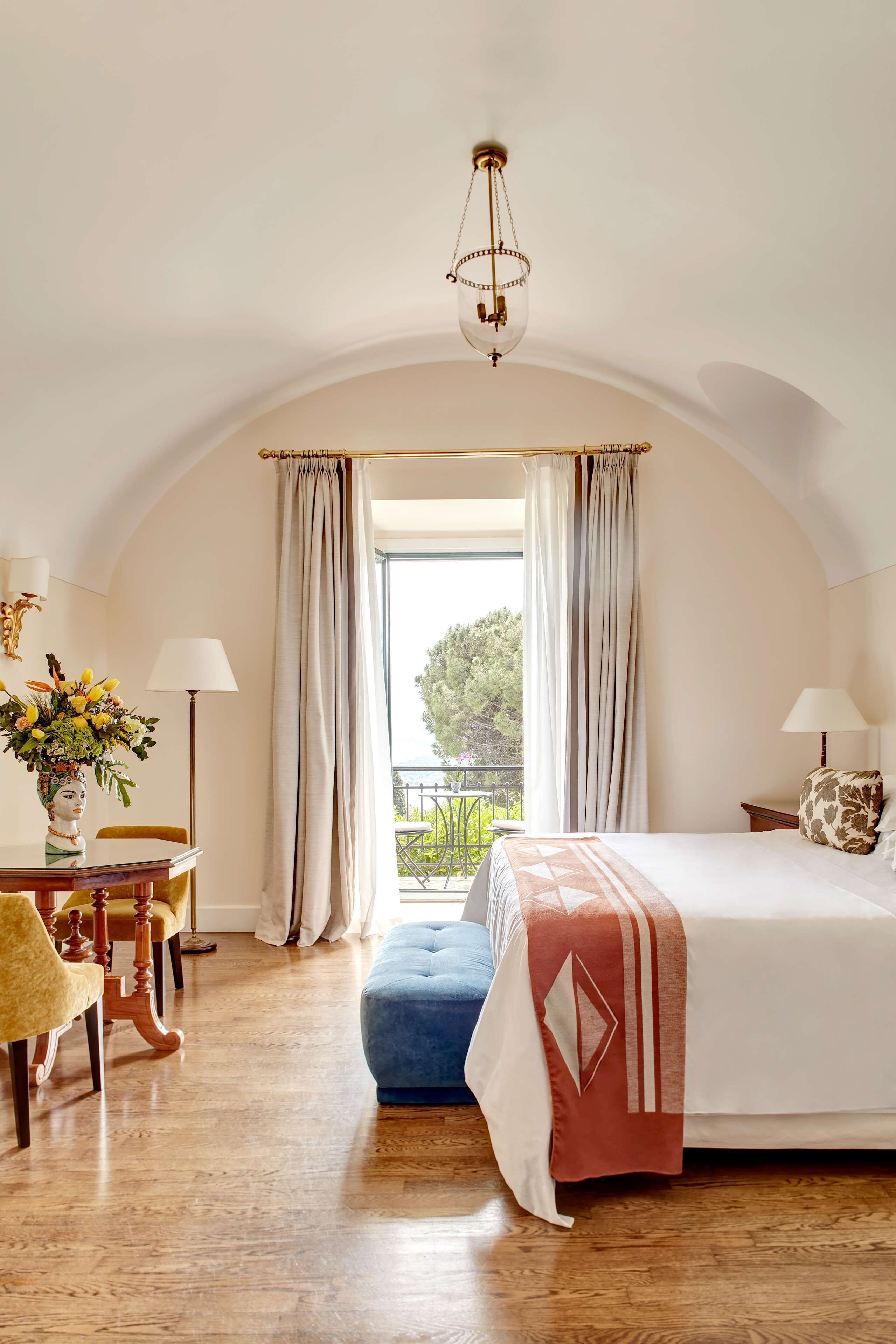 Quarto com cama grande, piso de madeira, varanda com cortinas e um candelabro.