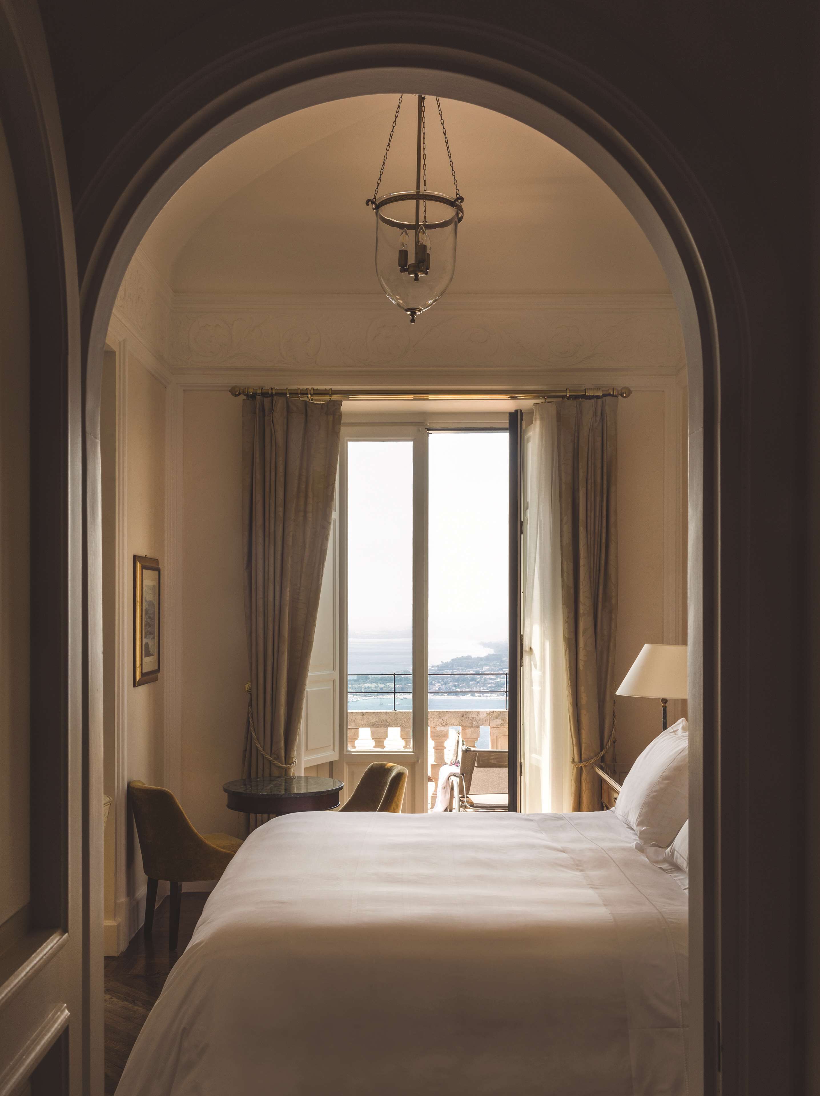 Quarto de hotel com cama, cadeira, escrivaninha e vista para o mar por uma janela grande.