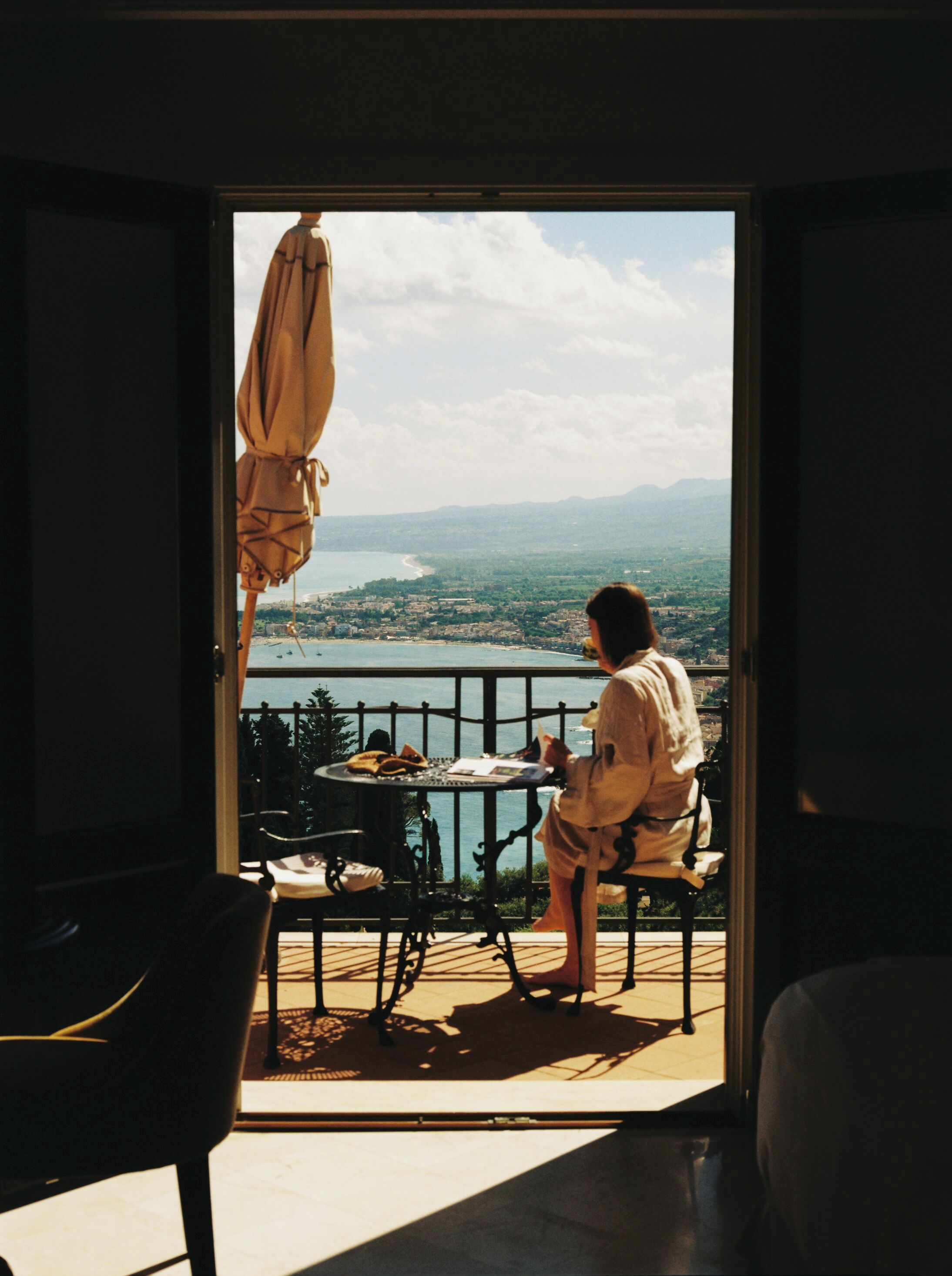 Pessoa sentada à mesa em varanda com vista para um corpo de água e paisagem distante.