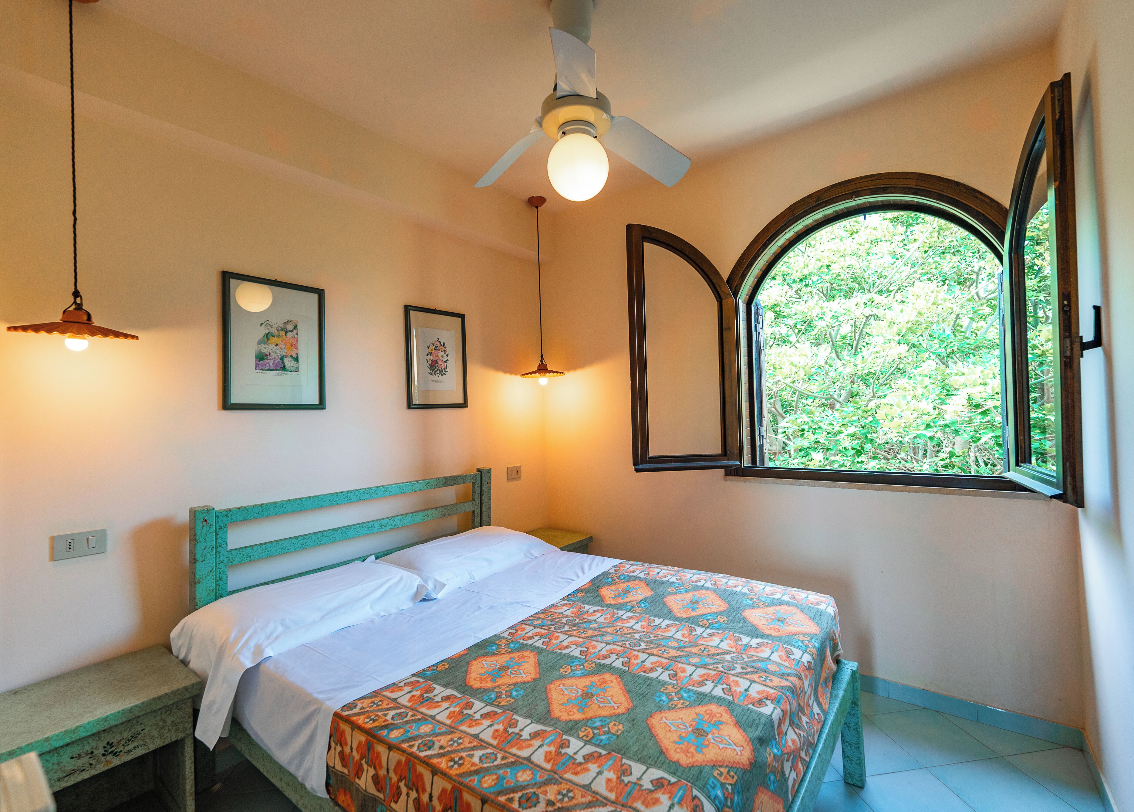 Quarto com cama, ventilador de teto, janela com persiana aberta e duas luzes de parede.