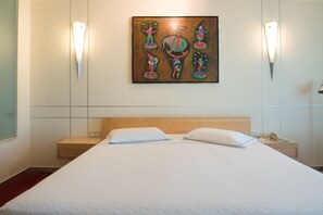 Quarto de hotel com uma cama grande, duas mesas de cabeceira, um criado-mudo, um quadro pendurado na parede e duas arandelas.