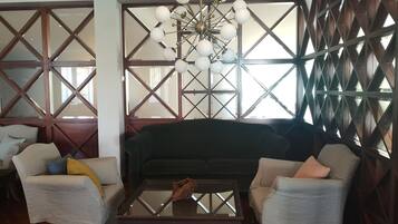 Uma sala de estar com um sofá verde escuro, duas poltronas brancas, uma mesa de centro de vidro e um lustre singular.