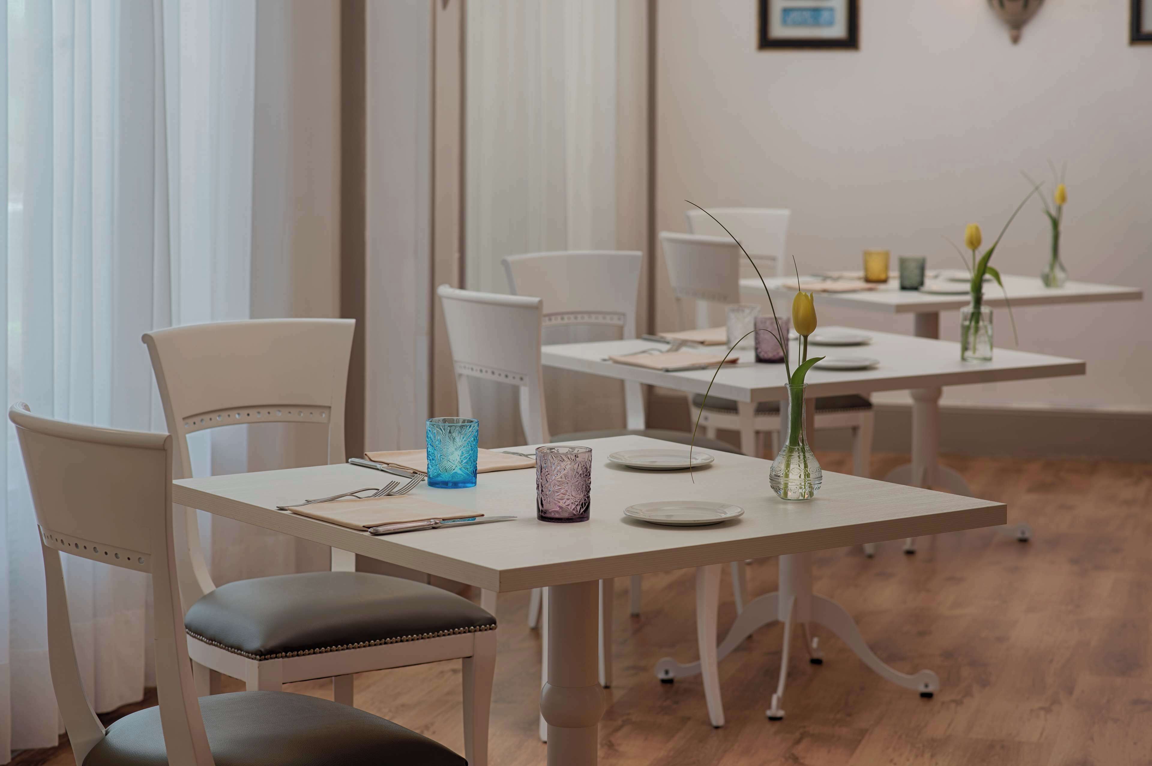 Uma sala de jantar com mesas e cadeiras brancas, castiçais de vidro e pequenos vasos com flores.