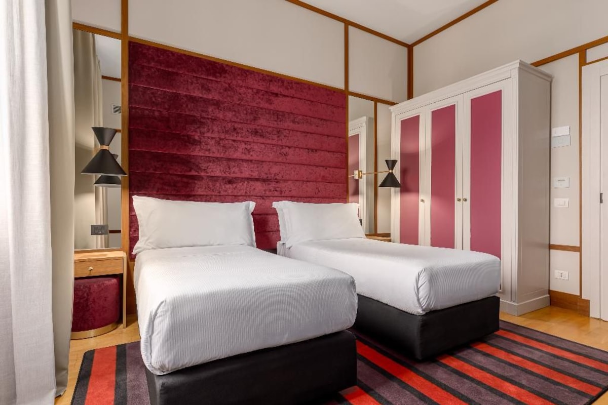 Quarto duplo com duas camas, uma parede de destaque vermelha, um guarda-roupa e um tapete listrado.