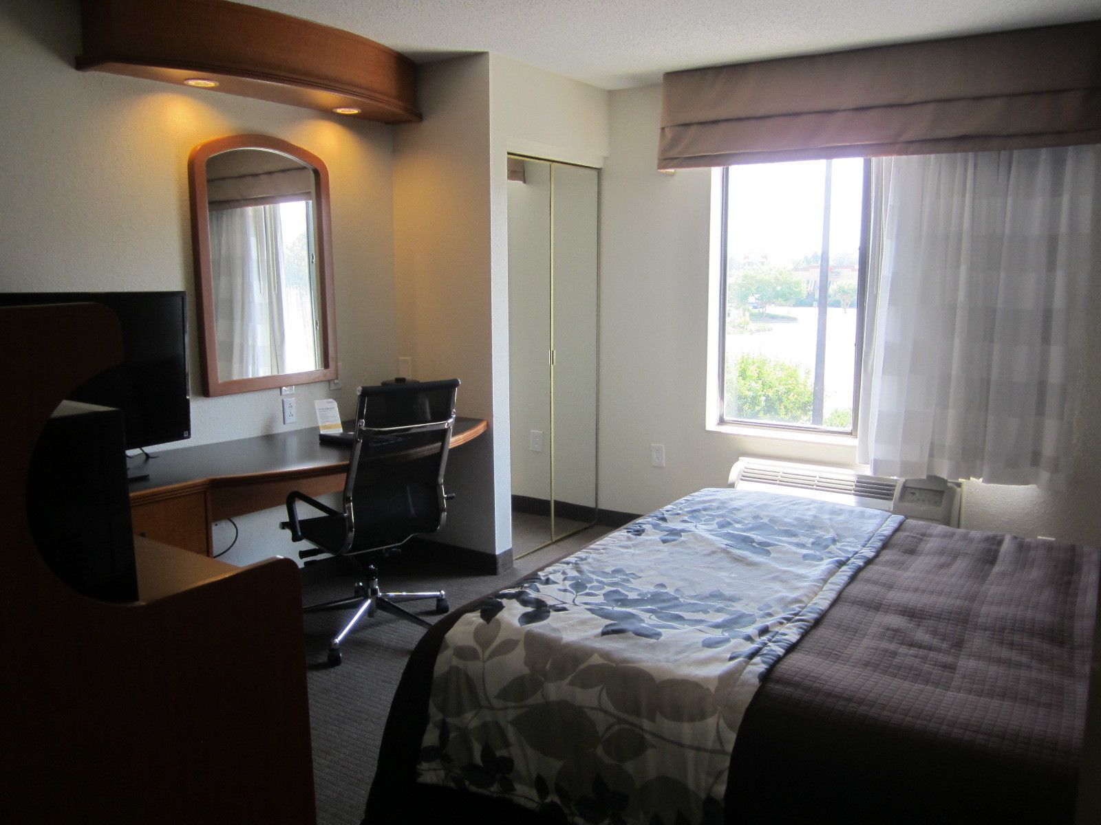 Quarto de hotel com cama, escrivaninha com cadeira, televisão, espelho e janela com cortinas.