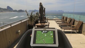 Convés de barco com uma tela de TV exibindo um jogo de futebol, cadeiras de lounge e vista do oceano e terra ao longe.