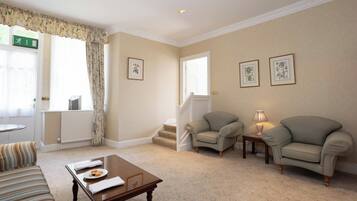 Quarto de hotel com sofá listrado, mesa de centro de madeira, duas poltronas, uma mesinha com abajur, escada e quadros na parede.