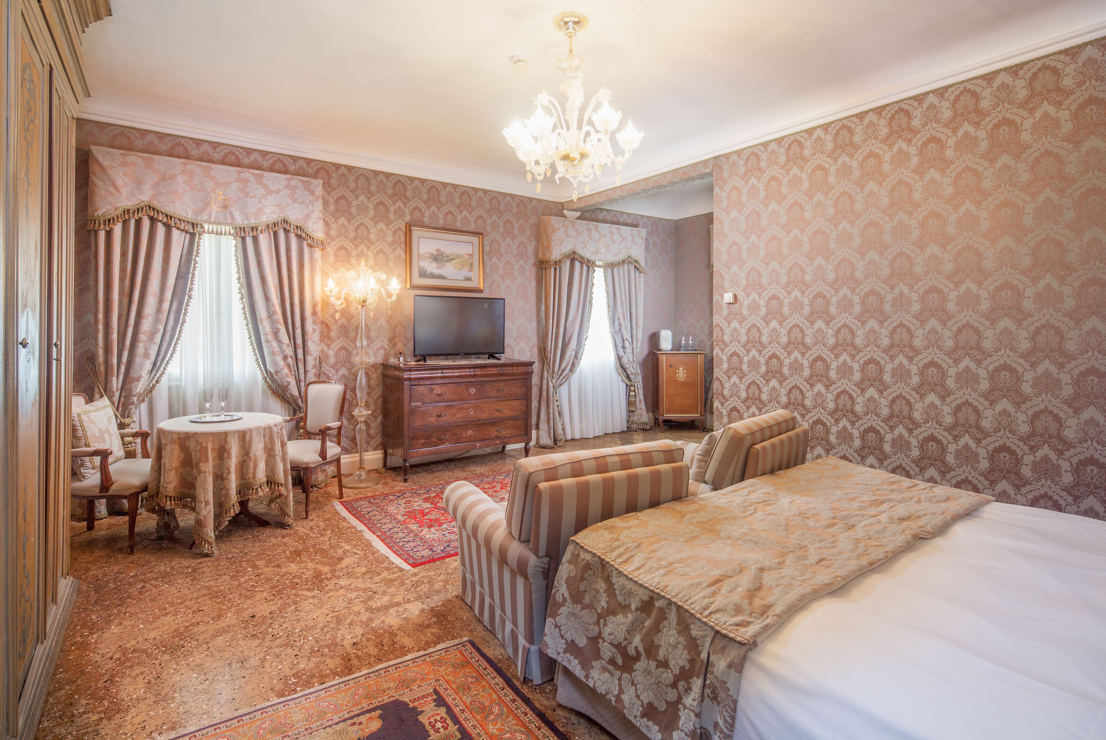 Quarto de hotel com uma cama, televisão, secretária com cadeira, pequena mesa e chão coberto de carpete.