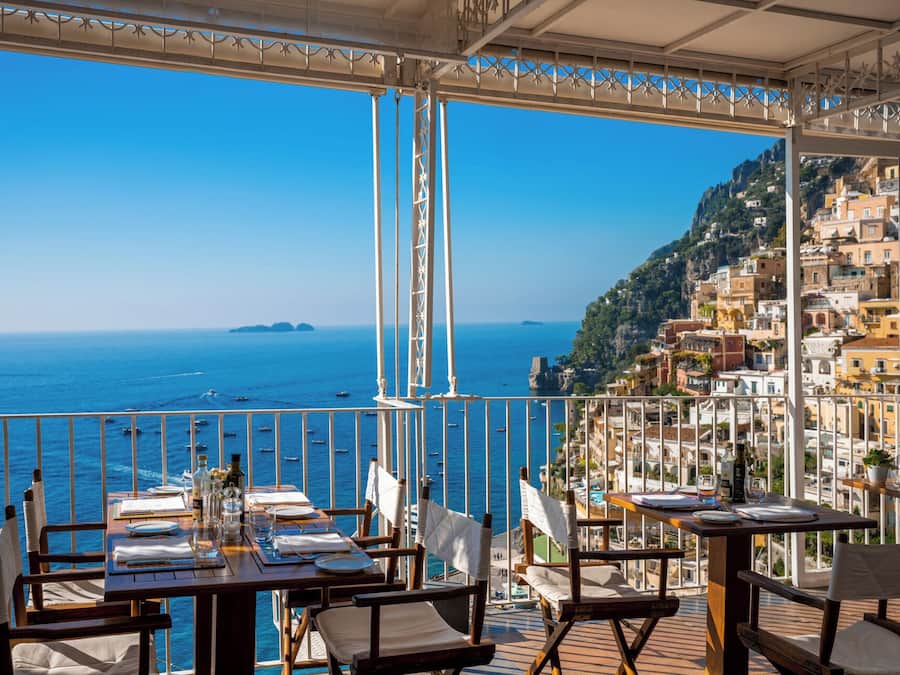 Terraço com mesas e cadeiras, com vista para uma vila costeira e o mar.