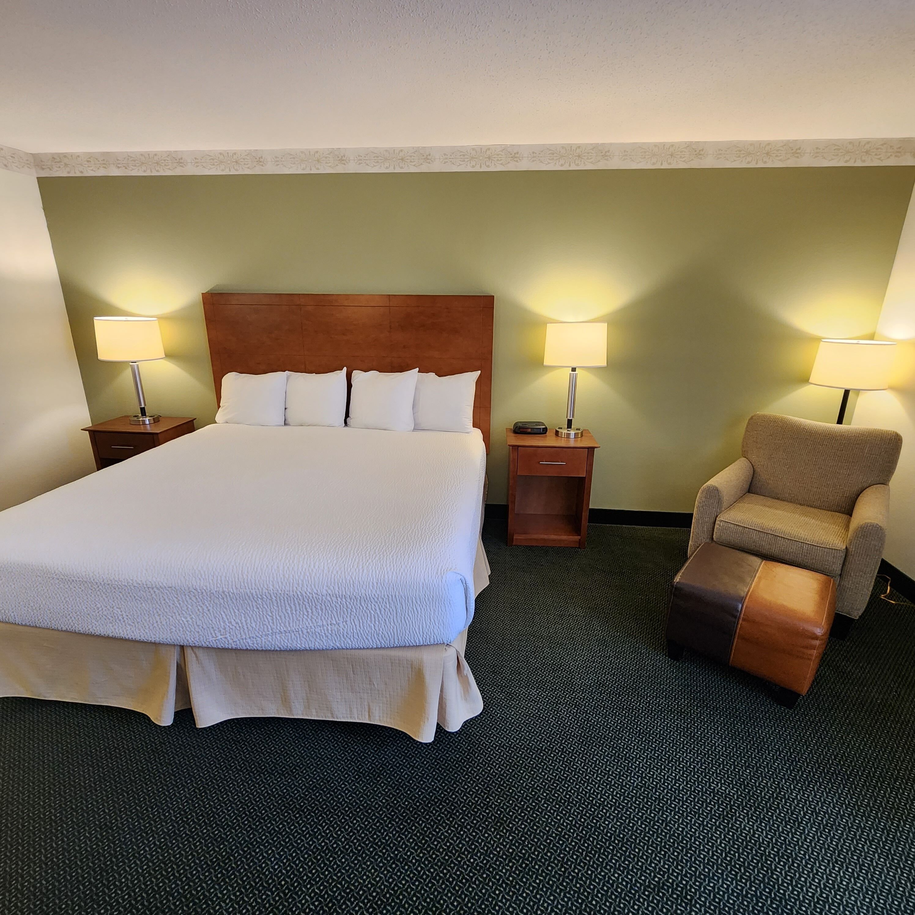 Quarto de hotel com uma cama grande, duas mesas de cabeceira com luminárias, uma cadeira e piso coberto por carpete.