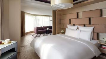 Quarto de hotel com uma cama grande, um sofá, uma escrivaninha com cadeira, uma luminária e vista para o exterior.
