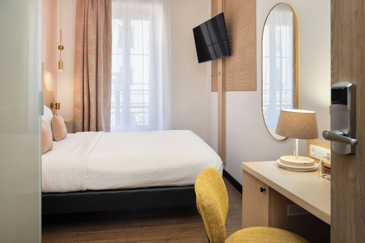 Quarto de hotel com cama, escrivaninha com abajur, televisão fixada na parede e um espelho grande.