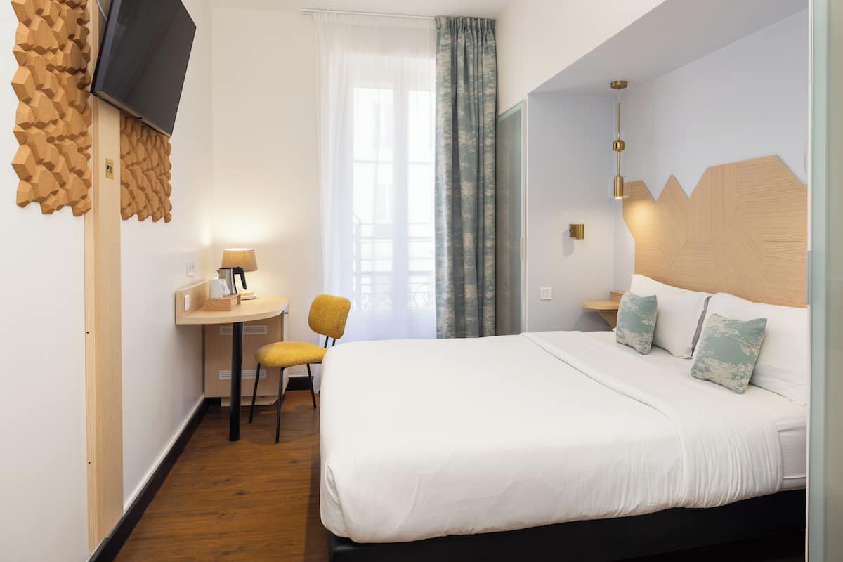 Quarto de hotel com cama, escrivaninha, cadeira, televisão e janela com cortinas.