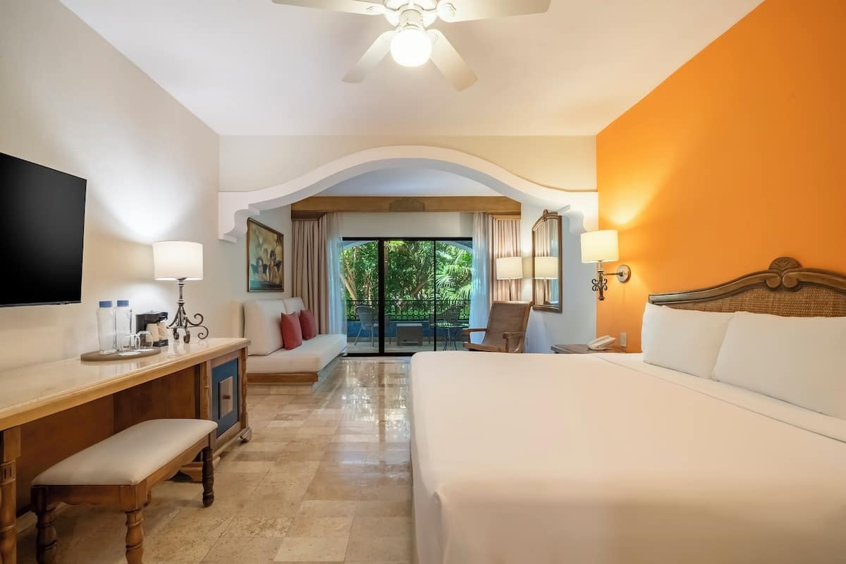 Quarto de hotel com uma cama grande, uma escrivaninha com um banco, uma televisão de tela plana, uma varanda com vista para o verde e um ventilador de teto.