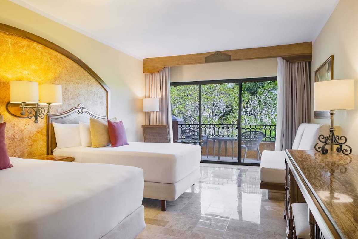Quarto de hotel com duas camas, varanda com vista para áreas verdes e sala de jantar com mesa e cadeiras.