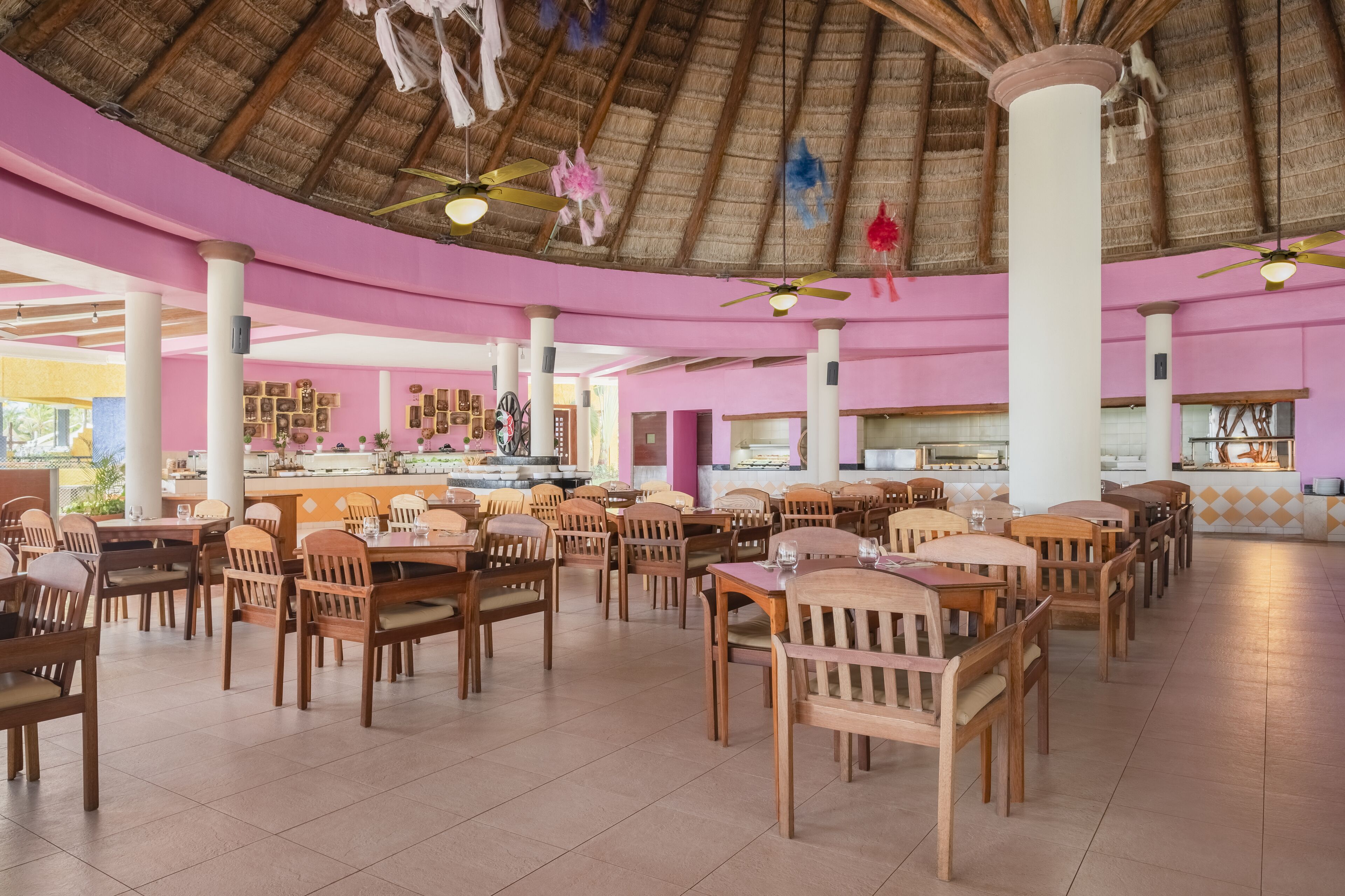 Uma sala de jantar ampla com mesas e cadeiras de madeira, telhado de palha e paredes cor-de-rosa.