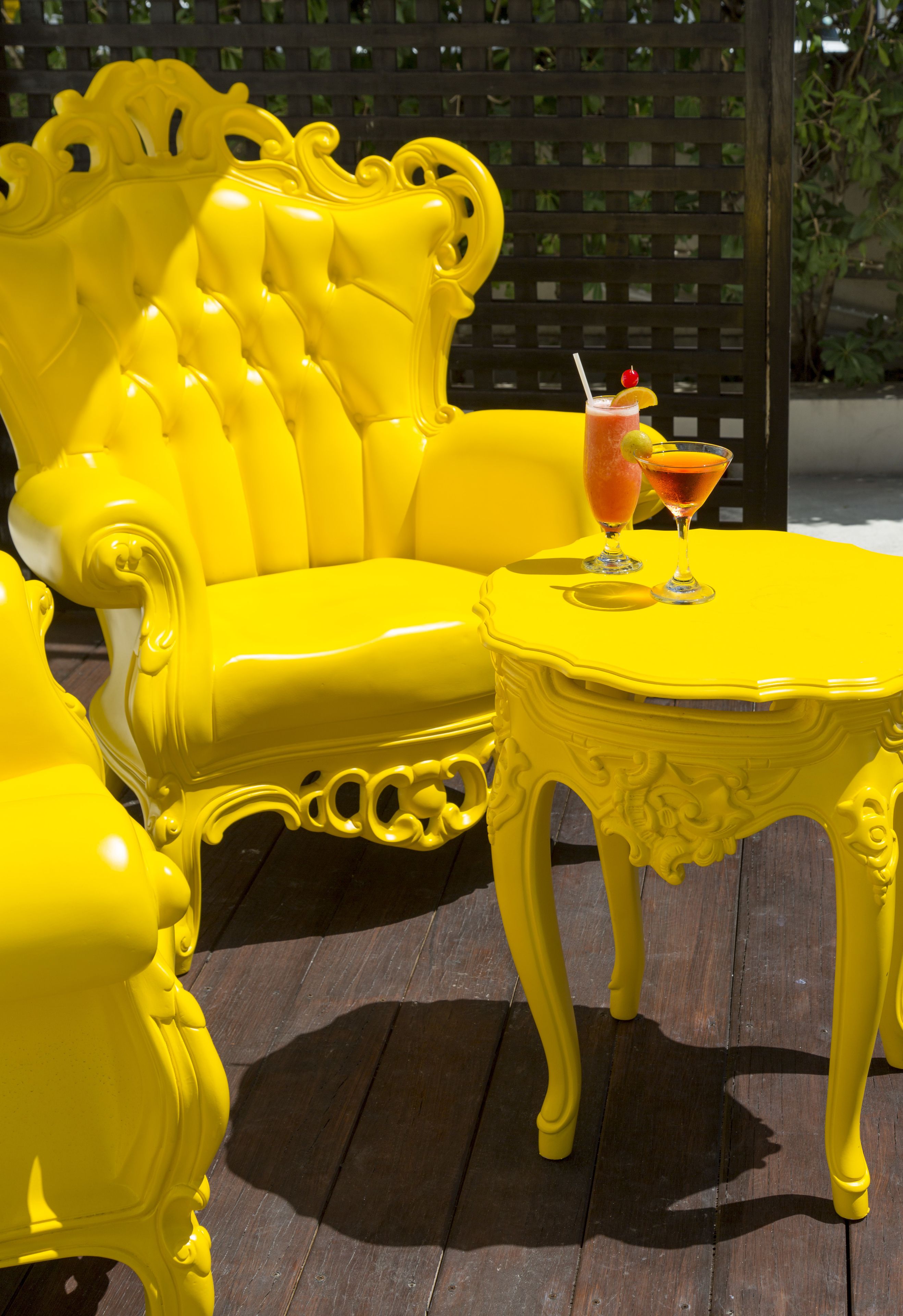 Conjunto de cadeira estofada amarela e mesa sobre um deck de madeira, com um fundo de treliça escura.