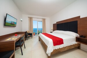 Quarto de hotel com cama, escrivaninha, cadeira, televisão e varanda com vista para o mar.