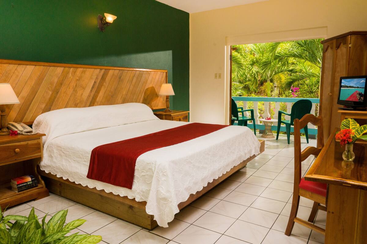 Quarto de hotel com cama, luminárias de cabeceira, mesa de cabeceira de madeira, escrivaninha com cadeira, televisão e vista para uma varanda com plantas.