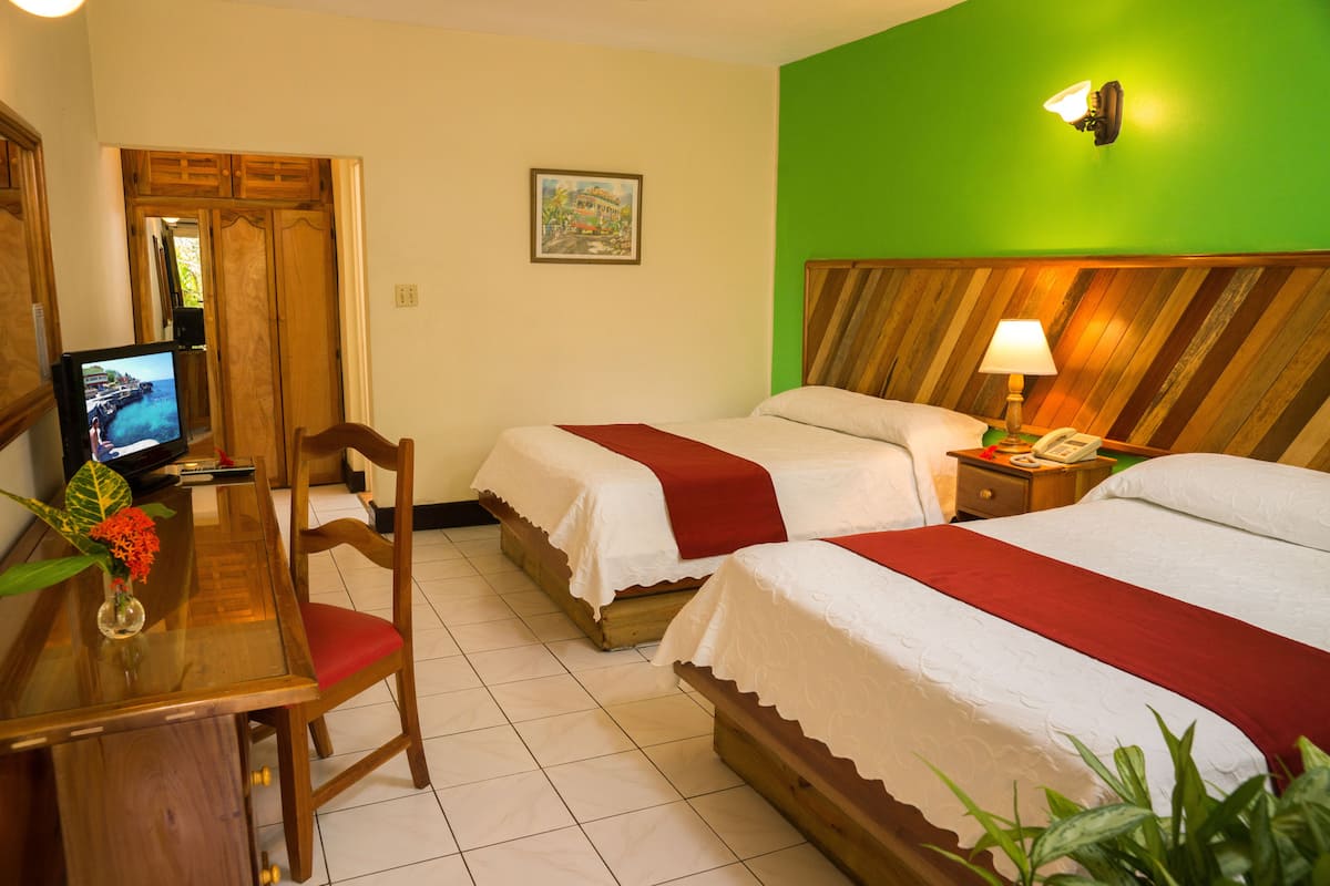 Quarto de hotel com duas camas, uma escrivaninha com computador, uma cadeira, televisão e parede verde.