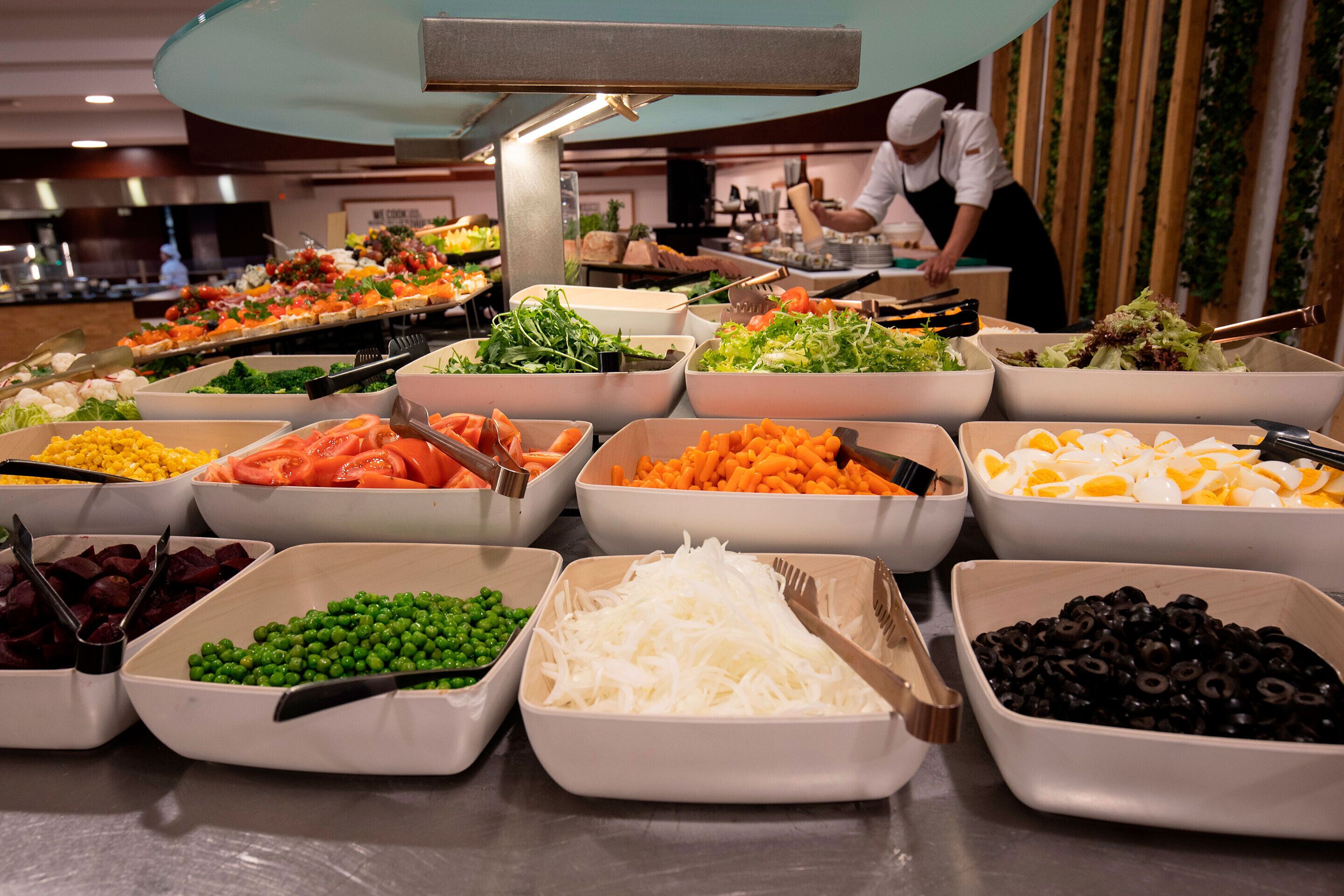 Buffet com diversos pratos em bandejas, incluindo legumes, massa e outras opções.