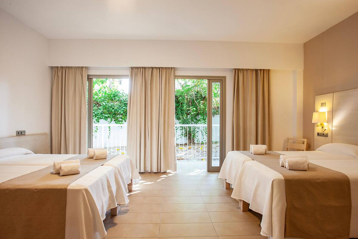 Quarto de hotel com duas camas, varanda com cortinas e vista para o verde.