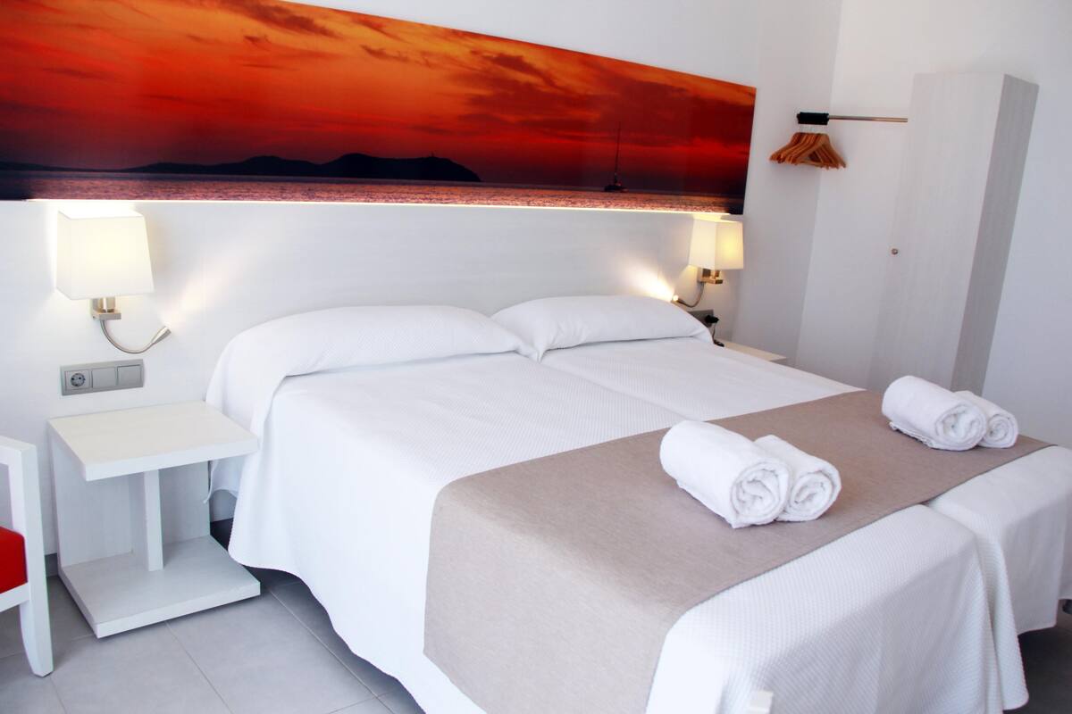 Quarto de hotel com uma cama grande, duas mesas de cabeceira, um criado-mudo e uma pintura de um pôr do sol sobre o mar.