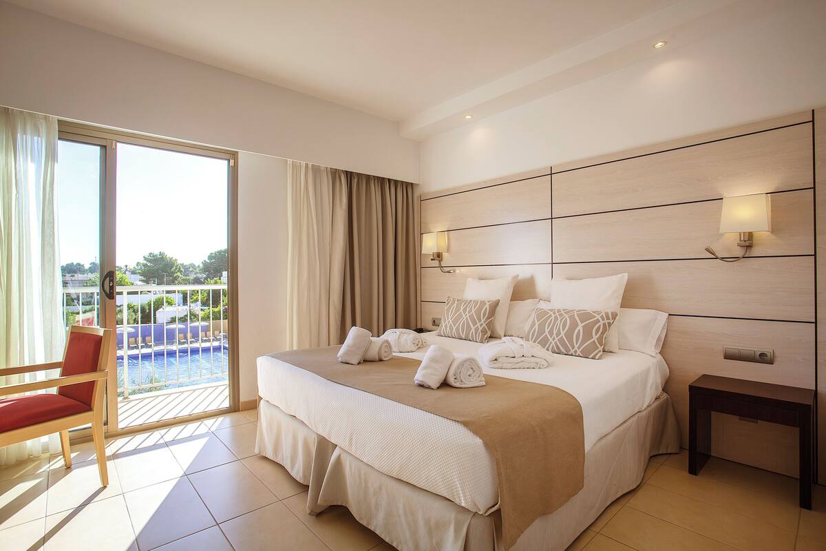 Quarto de hotel com uma cama grande, uma mesinha de cabeceira, uma cadeira e vista para uma piscina através de uma porta de correr.