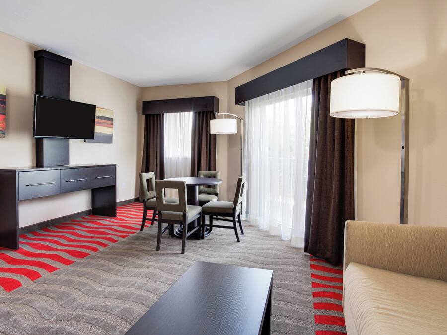 Quarto de hotel com televisão de tela plana, escrivaninha, mesa de jantar com cadeiras, sofá e uma grande janela com cortinas.