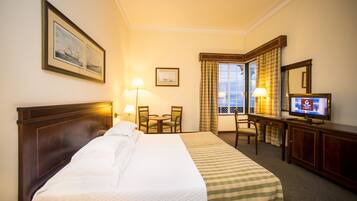 Quarto de hotel com uma cama grande, uma mesa de trabalho com televisão, duas cadeiras, uma janela com cortinas e obras de arte emolduradas na parede.