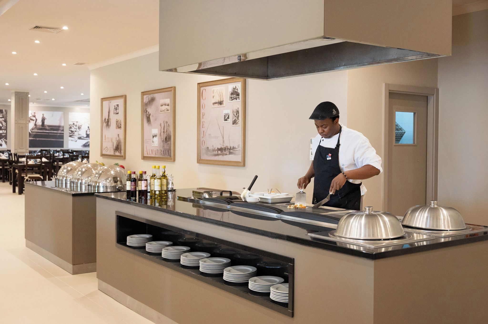 Buffet montado com um chefe preparando comida, balcão com pratos e acompanhamentos, e quadros nas paredes.