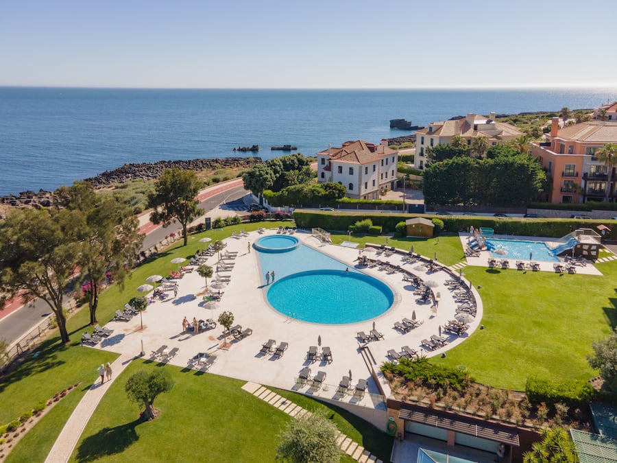 Um resort com uma grande piscina circular, uma piscina menor e uma área de praia. Há várias espreguiçadeiras e guarda-sóis ao redor das piscinas. O resort está localizado perto do oceano, com um litoral rochoso.