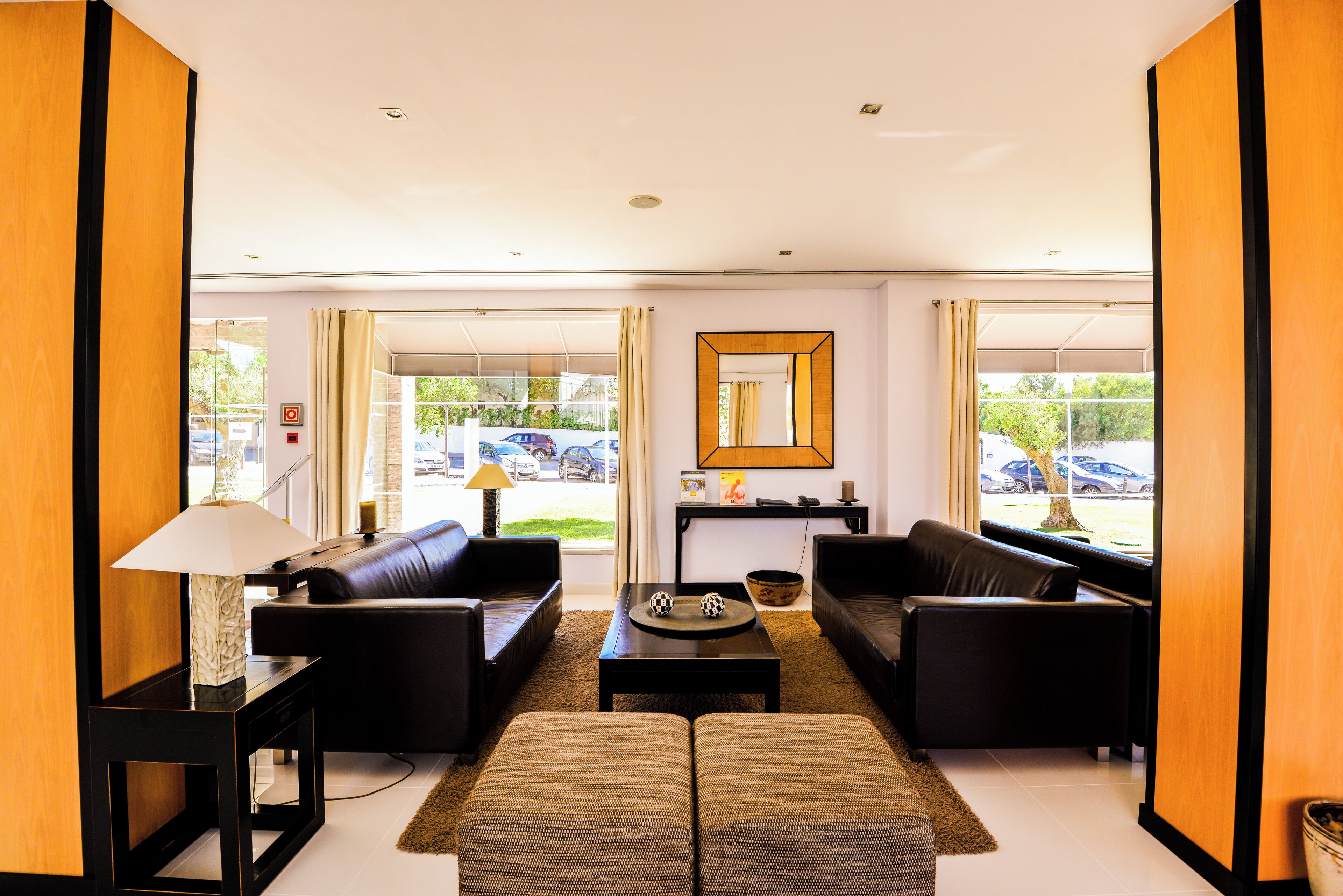 Quarto de hotel com um sofá, uma mesa de centro, uma luminária e vista para o estacionamento através de grandes janelas.