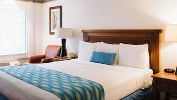 Quarto de hotel com uma cama grande, mesa de cabeceira com abajur, uma cadeira e relógio na parede.