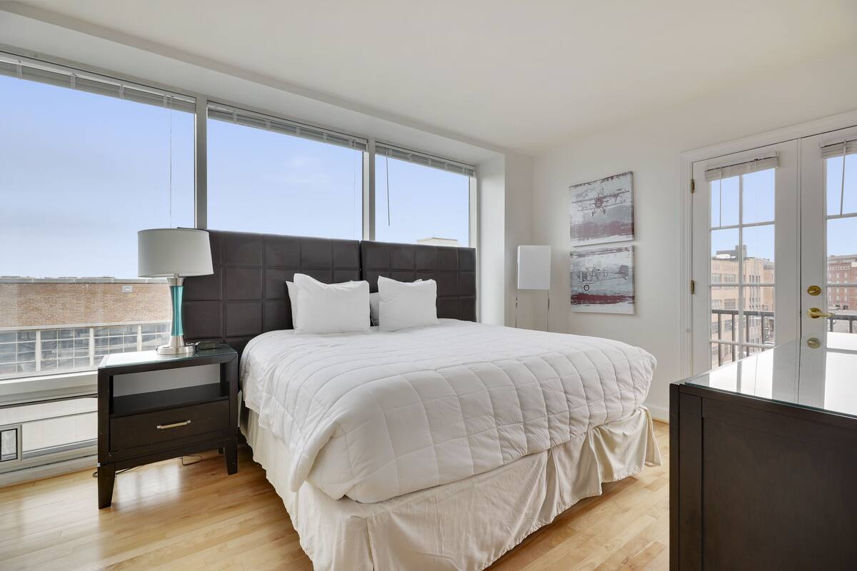 Quarto com cama grande, mesa de cabeceira, abajur e vista da cidade por grandes janelas.