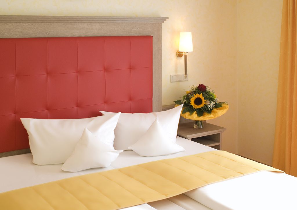Quarto de hotel com cabeceira vermelha, almofadas brancas, colcha amarela, mesa de cabeceira com abajur e um vaso com flores.