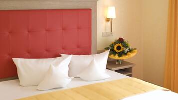 Quarto de hotel com cabeceira vermelha, almofadas brancas, colcha amarela, mesa de cabeceira com abajur e um vaso com flores.