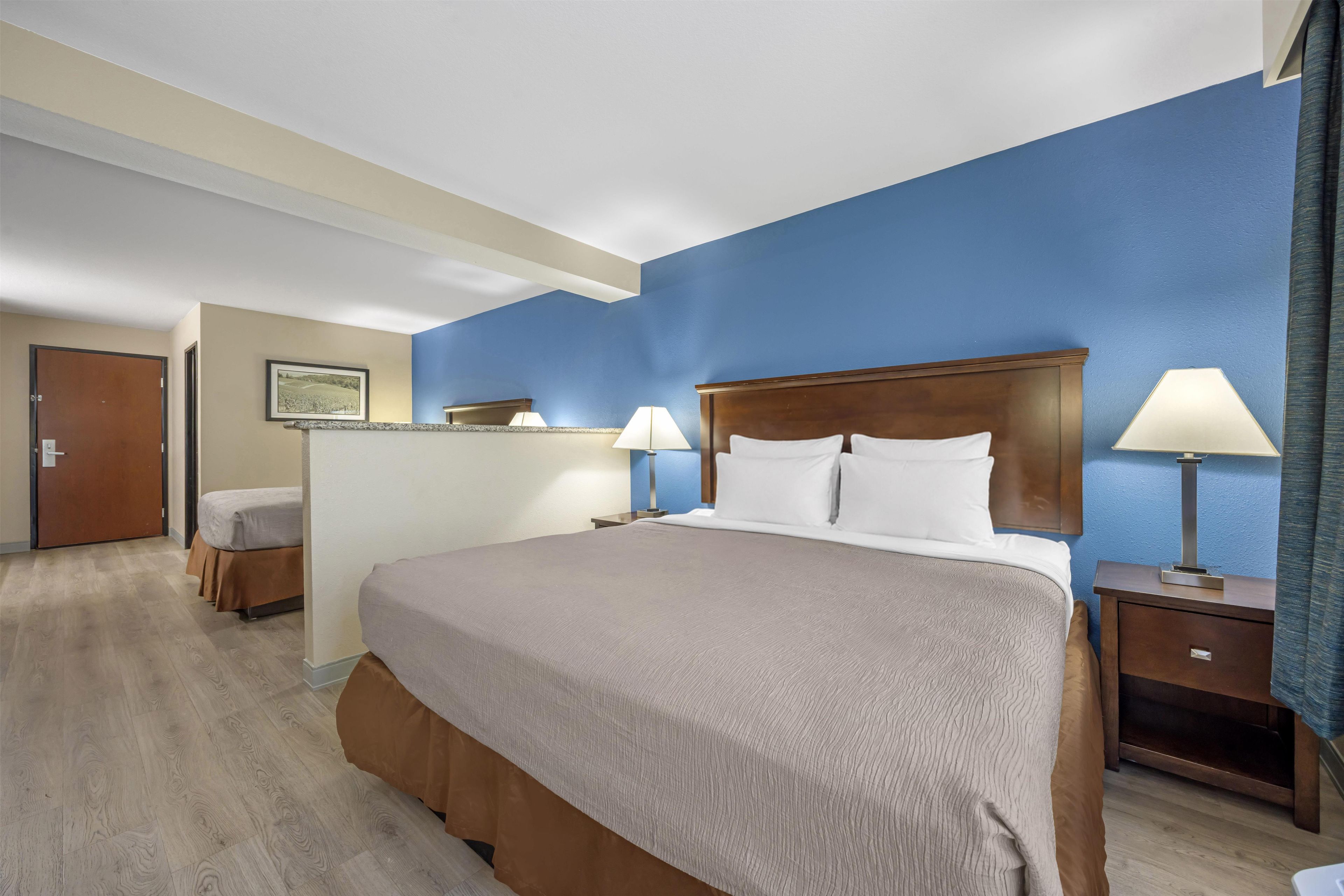 Quarto de hotel com uma cama grande, duas mesas de cabeceira com luminárias, um painel de cabeceira de madeira, uma porta e uma pequena escrivaninha com espelho.