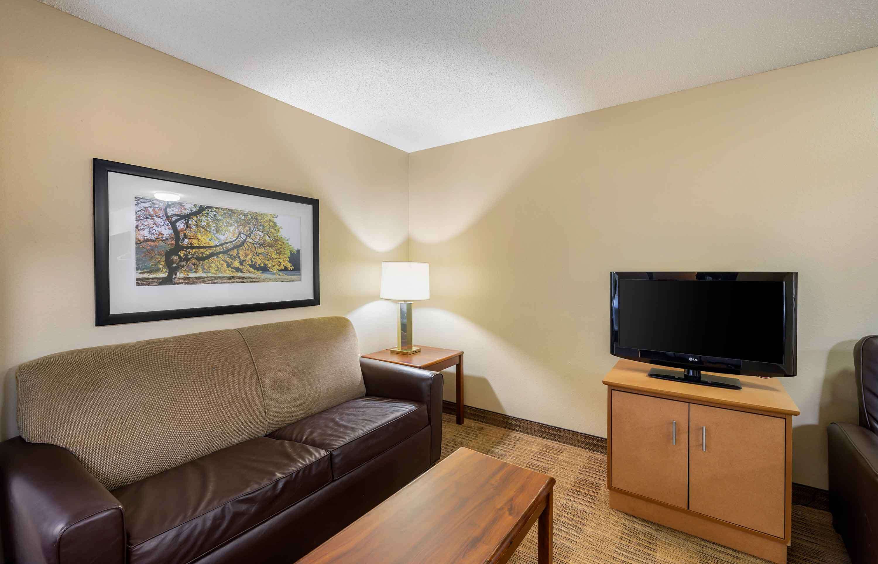 Quarto de hotel com sofá de couro marrom, mesa de centro de madeira, televisão sobre suporte de madeira, quadro emoldurado na parede e abajur em mesa lateral.