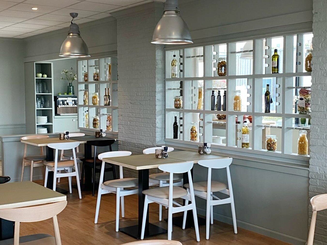 Interior de restaurante com cadeiras e mesas brancas, e uma parede de tijolos. Há prateleiras com garrafas e potes na parede.